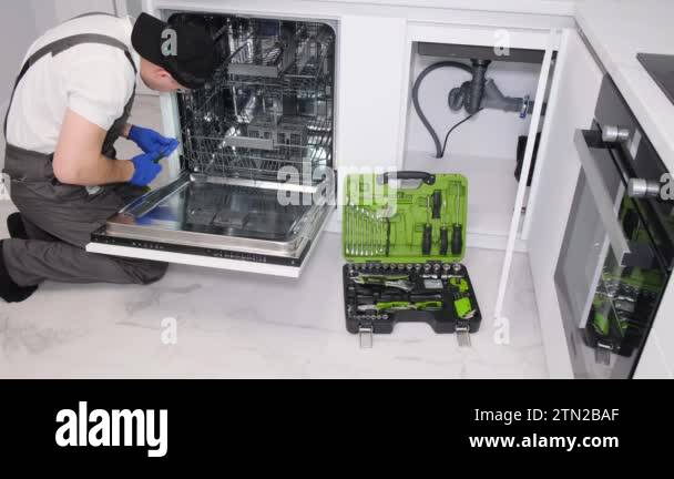 A professional repairman repairs a broken dishwasher in a modern kitchen. Mechanical breakdown ...