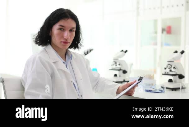 Male scientist with long hair holds clipboard sitting at table with ...