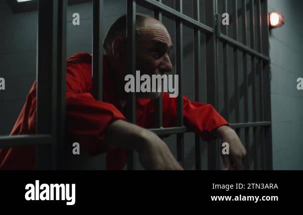 Elderly prisoner in orange uniform stretches fingers, leans on metal ...