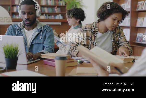 Diverse group of students doing assignments in library reading books and using laptops sitting ...