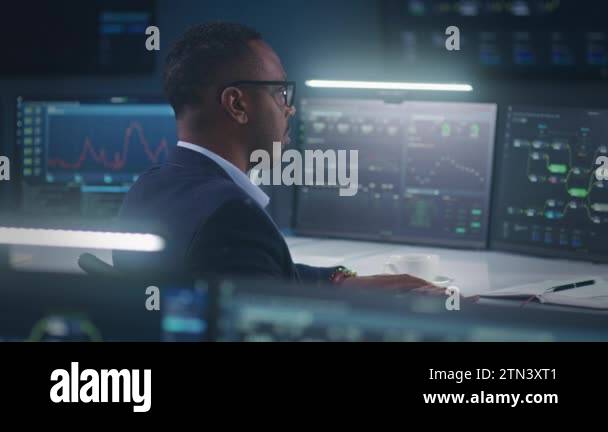 African American software engineer sits in front of computer and big digital screens with data ...