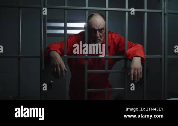 Elderly prisoner in orange uniform holds hands on metal bars, looks at ...