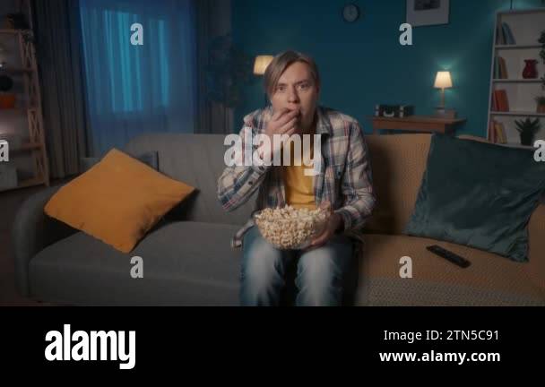 Horror. Shocked man watching a scary movie on TV at home and eating ...
