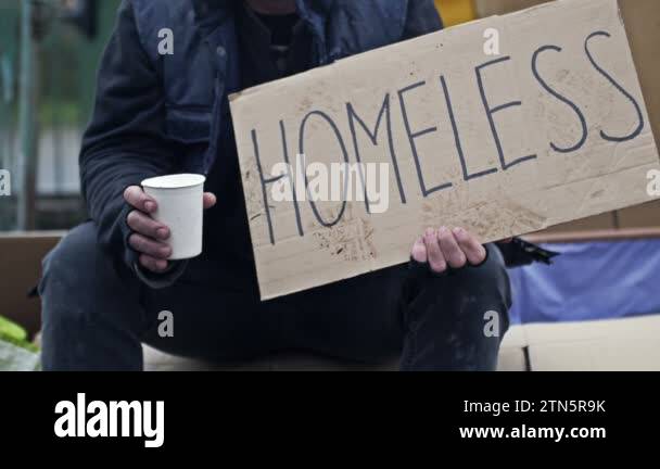 Untidy man sits with an alms cup and holds a handwritten HOMELESS ...
