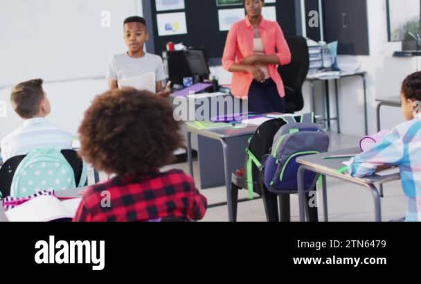 Diverse female teacher and happy schoolchildren at desks reciting in ...