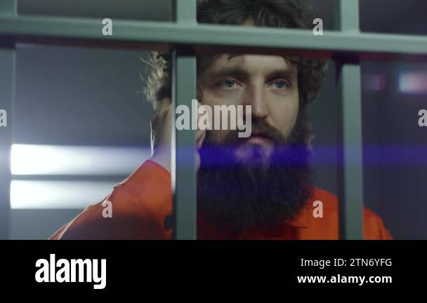 Prisoner in orange uniform stands behind metal bars, talks on phone in ...