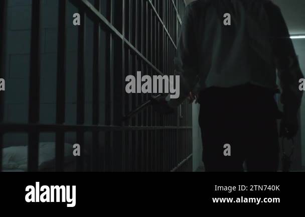 Prison employee with police baton watches criminals in prison cells. Prisoners in orange ...