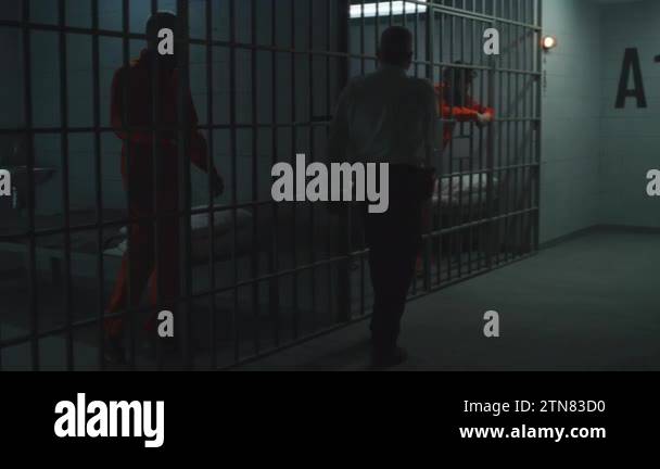 Prison officer with police baton watches criminals in prison cells ...