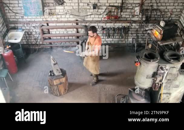 Beard man blacksmith in forge makes metal tools on iron anvil with ...