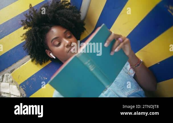Closeup teen reading book aloud lying on colorful bench outdoors. Curly ...
