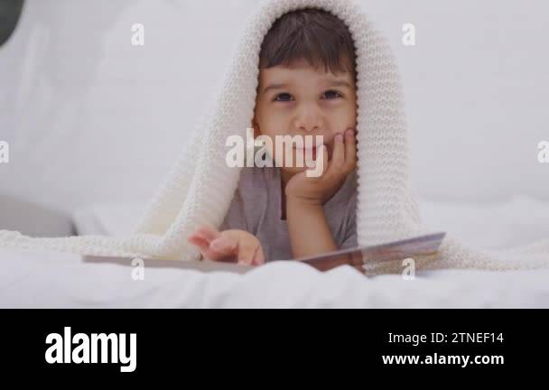 Cute smiling child boy looking into a book. Preschool kid lying on bed ...