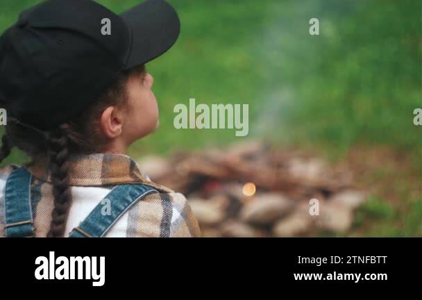 Selective focus of preteen girl in cap sitting near bonfire and look at fire. Little child lit ...