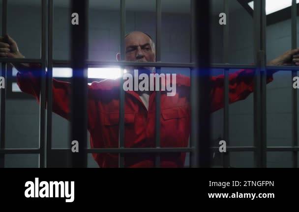 Elderly prisoner in orange uniform holds hands on metal bars in jail ...