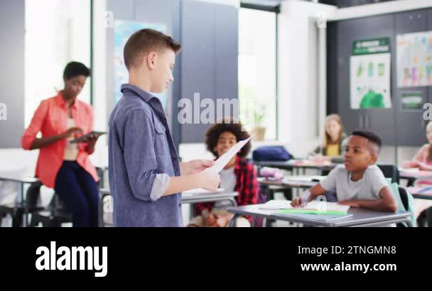Diverse female teacher and happy schoolchildren at desks reciting in ...