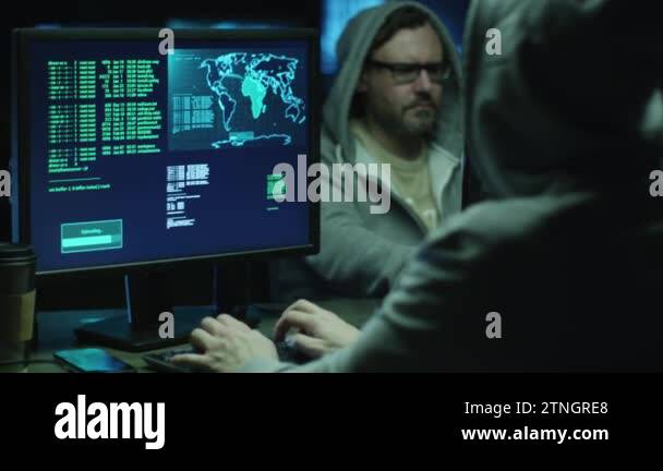 Two hackers in hoods work on a computers with maps and data on display ...