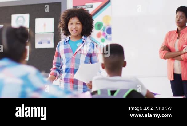 Diverse female teacher and happy schoolchildren at desks reciting in ...