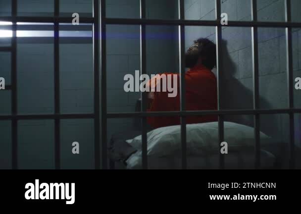 Prisoner in orange uniform sits on the bed in prison cell with barred window. Guilty criminal ...