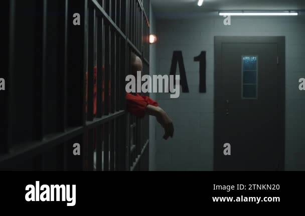 Elderly prisoner in orange uniform stretches his hands, leans on metal ...