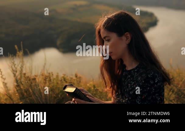 A beautiful woman sits on a hill above the river, admires the view at ...