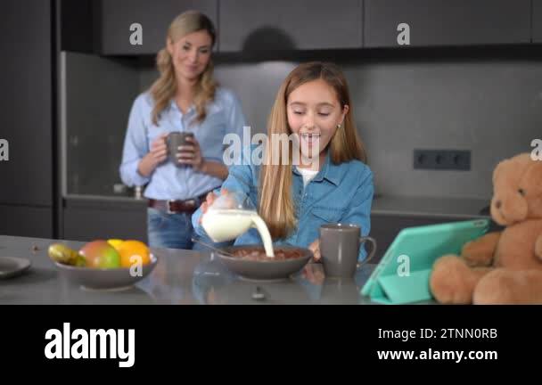 Smiling happy cute teen girl pouring milk in bowl with breakfast cereal as blurred woman talking ...