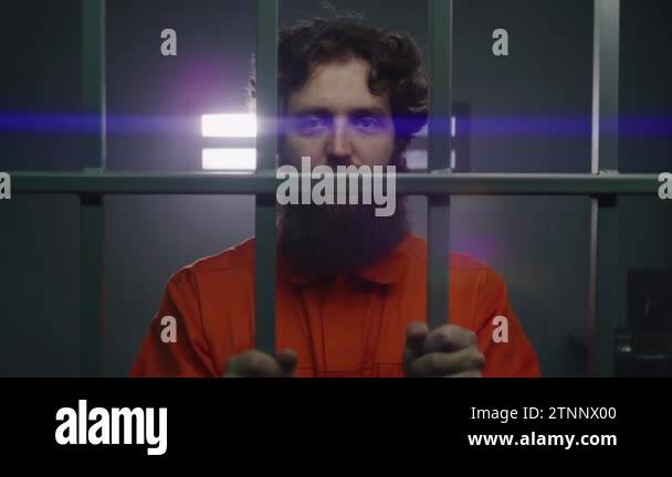 Prisoner in orange uniform holds metal bars, stands in prison cell in ...