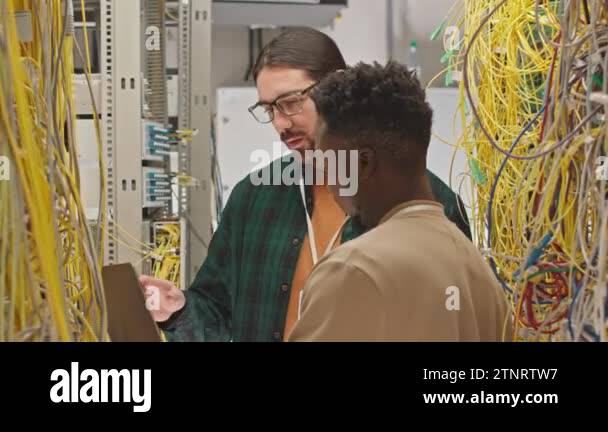 Two multiethnic male server room employees standing in narrow rack ...