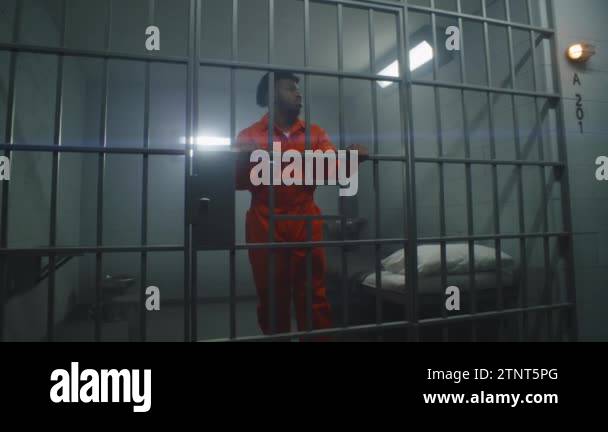 African American prisoner in orange uniform leans on jail cell bars and ...