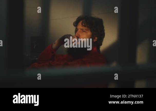 Depressed prisoner in orange uniform sits on bed, smokes cigarette in ...