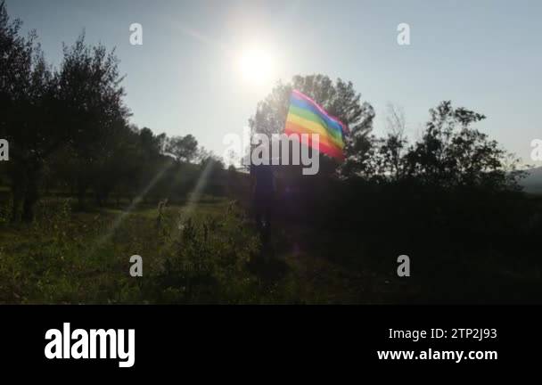Young woman alone with LGBTQ+ flag walking as it waves in the sunset ...