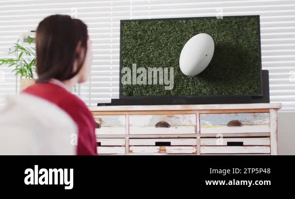 Caucasian woman watching tv with rugby ball at stadium on screen ...