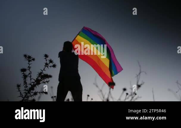 Young woman alone with LGBTQ+ flag walking as it waves in the sunset ...