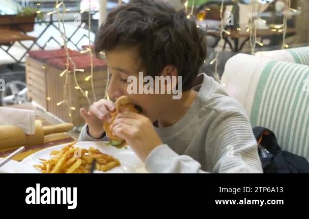 the boy is eating a hamburger in a cafe. 4k. a hamburger with a cutlet ...