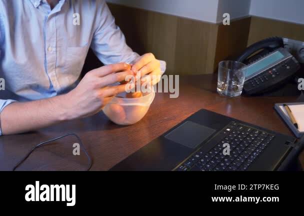 A man eats boiled chicken eggs from a food container, working on a laptop, late at night ...
