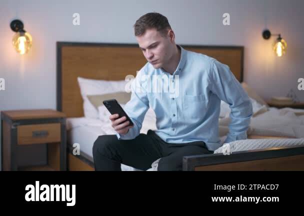 Serious young man sitting on bed scrolling smartphone touchscreen in slow motion. Portrait of ...