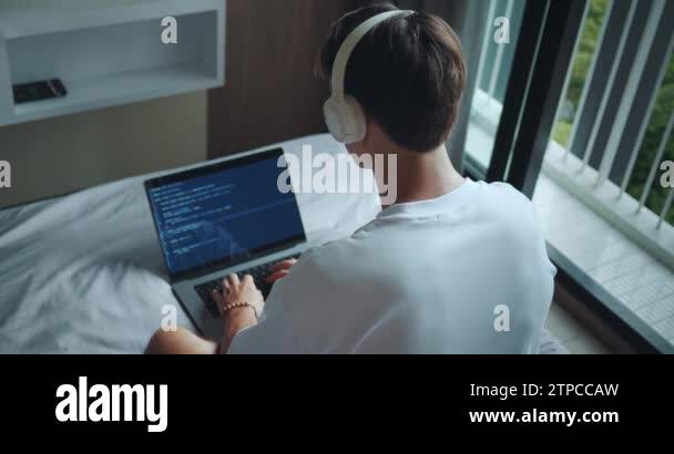 Rear view man freelancer programmer in headphones sitting on bed in bedroom and working remotely ...