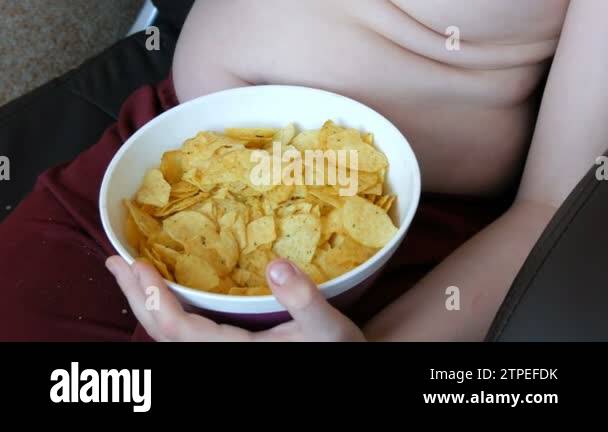 Fat teenager with fat folds on his body is eating potato chips ...
