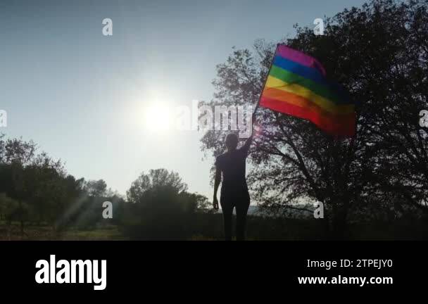 Young woman alone with LGBTQ+ flag walking as it waves in the sunset ...