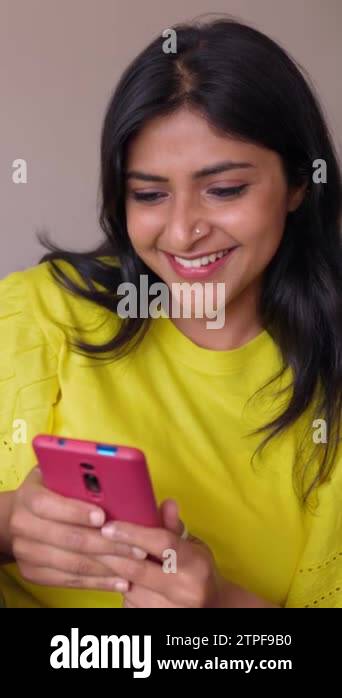 Happy young indian woman having fun using cell phone while sitting on couch at home. Cheerful ...