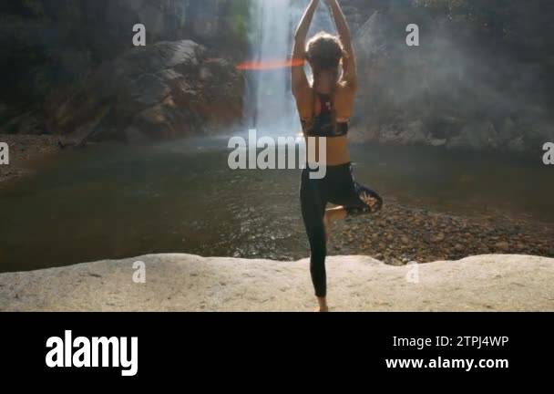 backside view girl stands in yoga pose Vrikshasana against waterfall ...