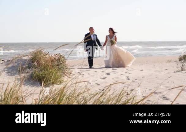 Lovely bride and groom holding hands, walking at sea coast. Attractive man in suit, woman in ...
