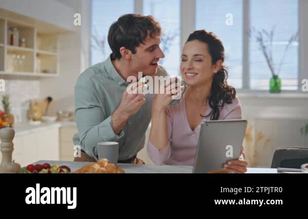 Laughing pair enjoy breakfast standing cozy kitchen with digital tablet ...