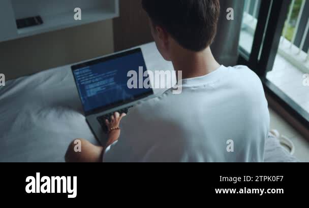 Rear view of Man Crafting Software Code on Laptop while sitting on bed at home. Freelance man ...