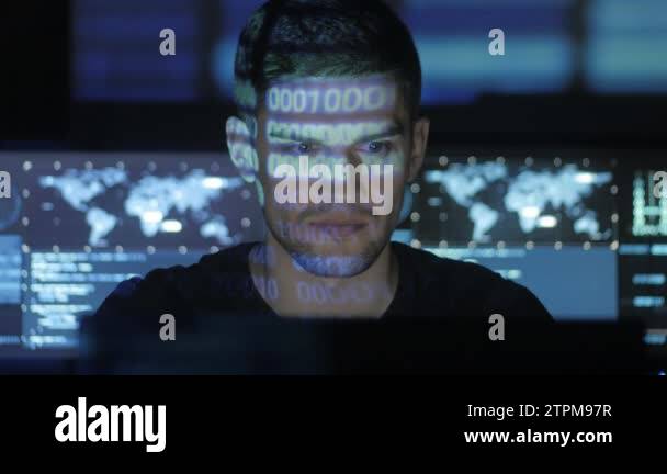 Male hacker programmer working at computer while blue code characters reflect on his face in ...