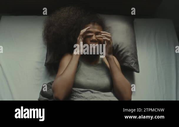 Top down shot of black teen girl lying in her bed, rubbing eyes and ...