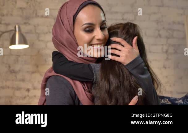 Young beautiful mother in hijab hugs her little daughter, looks at camera, sitting on couch ...