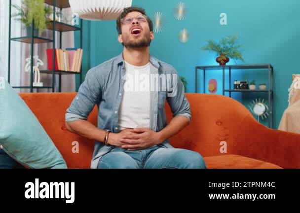Indian young man sits on sofa feeling sudden strong abdominal stomach ...