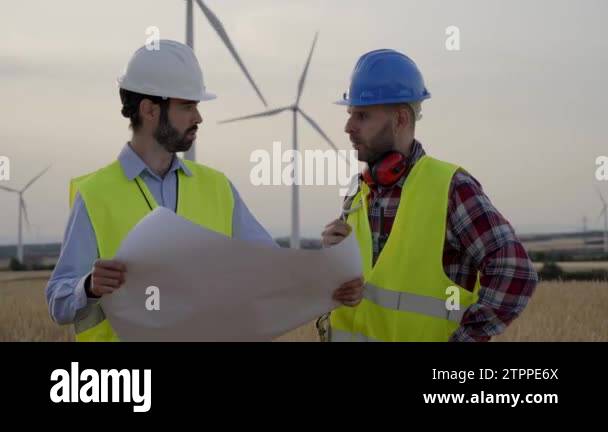 Two wind engineers in vests and hard hats look at plans of a wind ...
