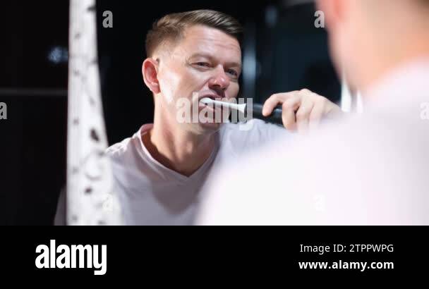 Focused handsome young man electric brushing teeth in front of mirror ...