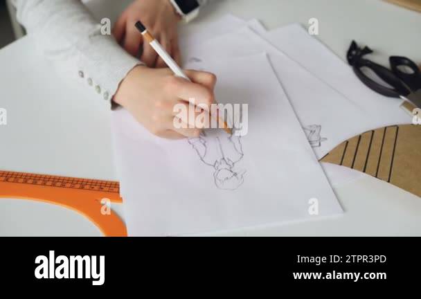 Close-up shot of womans hand drawing outlines of stylish ladies garment. Paper, tailors scissors ...