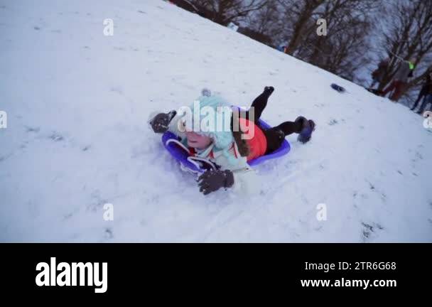 Little girl is lying on her belly on a sled, going down a hill in the ...